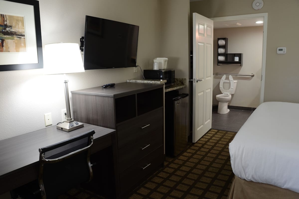 Quarto de hotel com escrivaninha, cadeira, cama, televisão, micro-ondas e banheiro com vaso sanitário e pia.