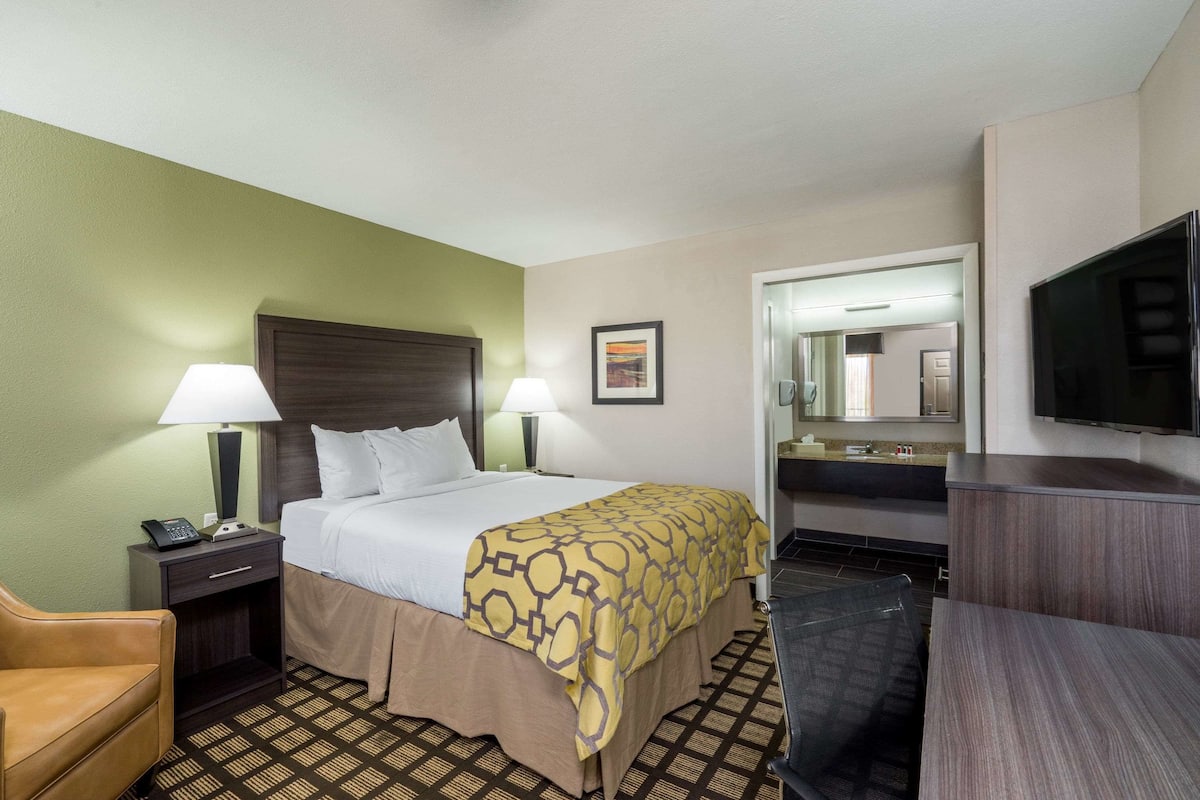 Quarto de hotel com cama, duas luminárias de cabeceira, escrivaninha com telefone, cadeira, televisão e espelho de banheiro.