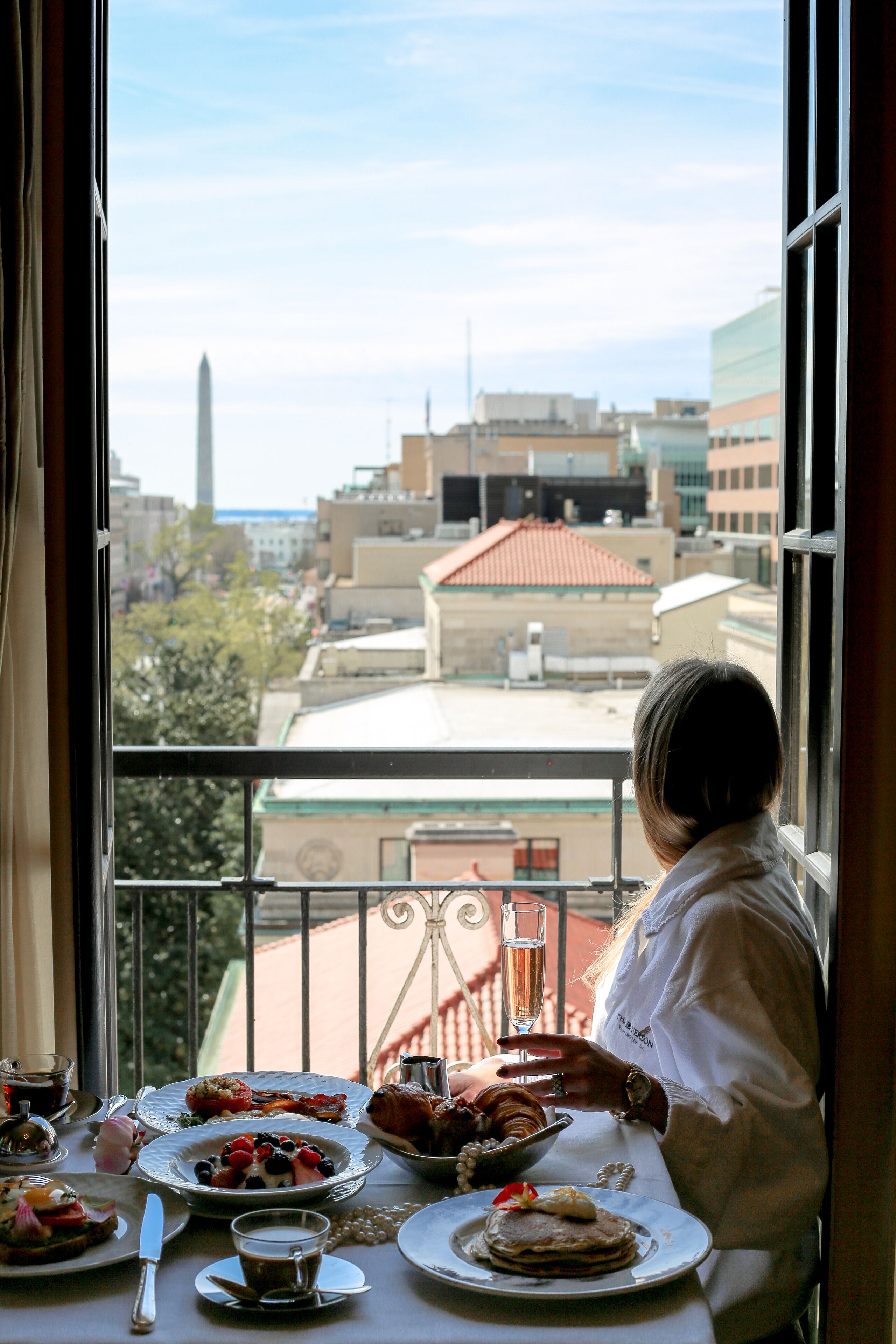 Mesa de jantar com diversos pratos e um copo de água, vista de uma varanda com vista para um horizonte urbano com edifícios e uma torre.