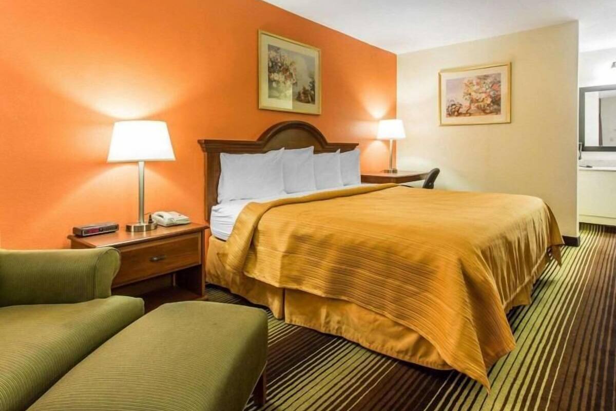 Quarto de hotel com cama, duas luminárias de cabeceira, mesa de cabeceira, uma cadeira e janela.