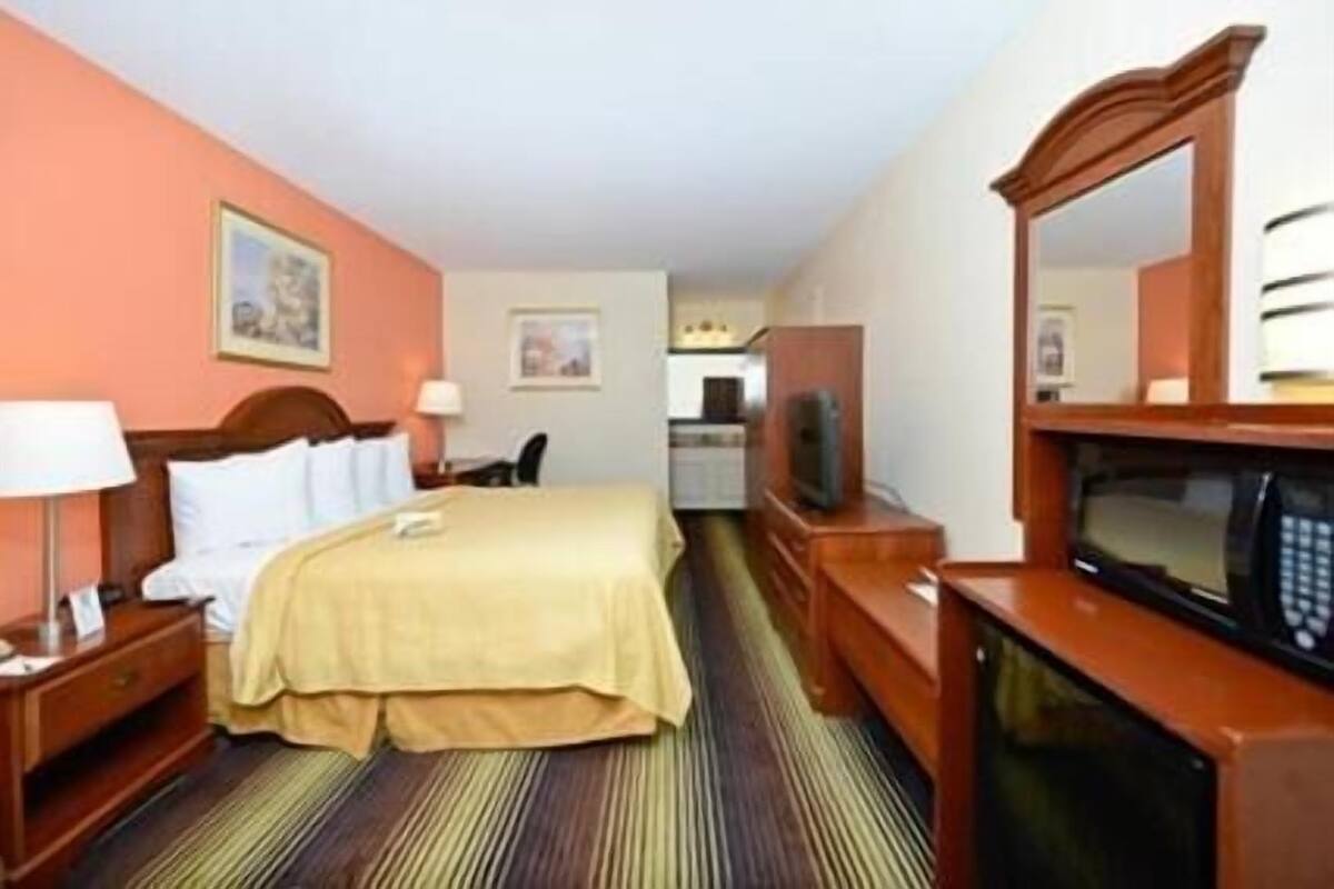 Quarto de hotel com cama, mesa de cabeceira, luminária, televisão e micro-ondas.
