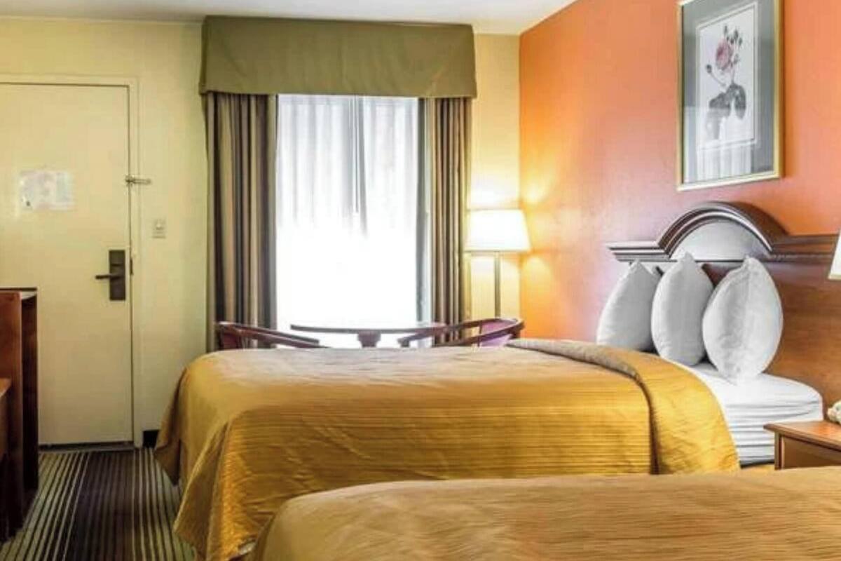 Quarto de hotel com duas camas, uma escrivaninha, uma luminária e um quadro na parede.
