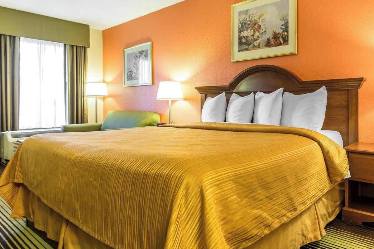 Quarto de hotel com uma cama grande, duas luminárias de cabeceira, uma cadeira verde e um quadro na parede.