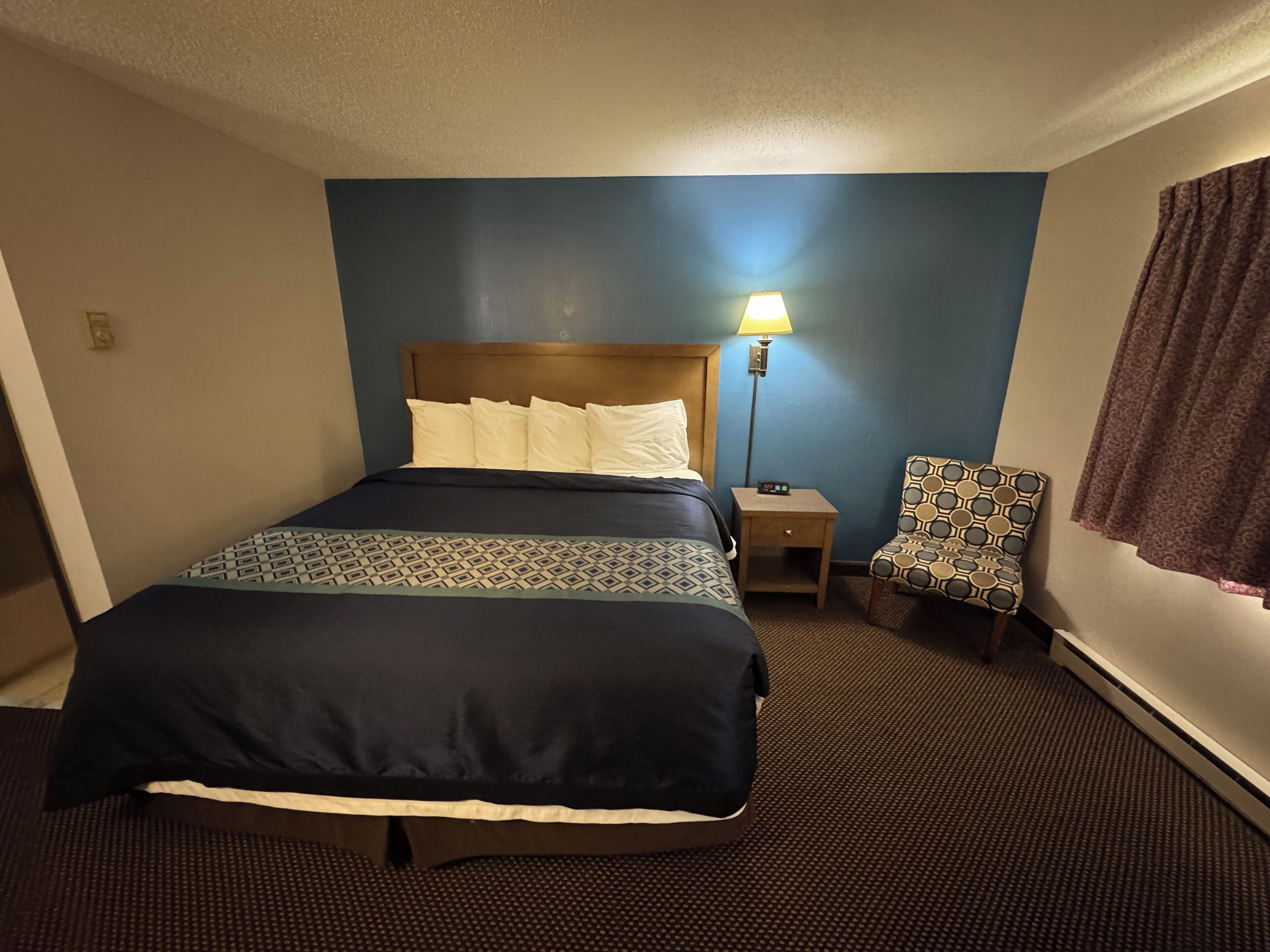 Quarto de hotel com uma cama grande, uma mesa de cabeceira com abajur, uma cadeira com almofada estampada e uma parede com detalhe azul.