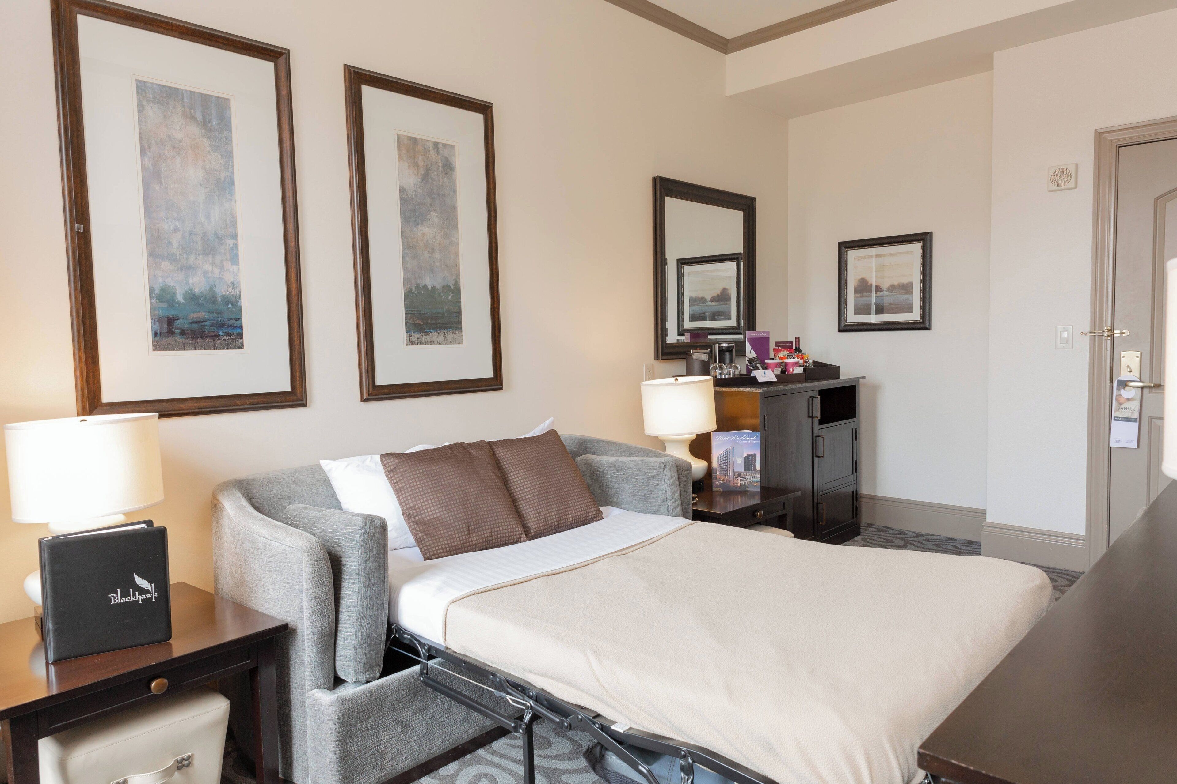 Quarto de hotel com cama, escrivaninha, cadeira, luminária e quadros na parede.