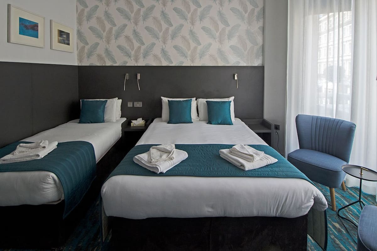 Quarto de hotel com duas camas, cada uma com colcha azul-esverdeada e roupa de cama branca. Há uma cadeira e uma pequena mesa no canto.