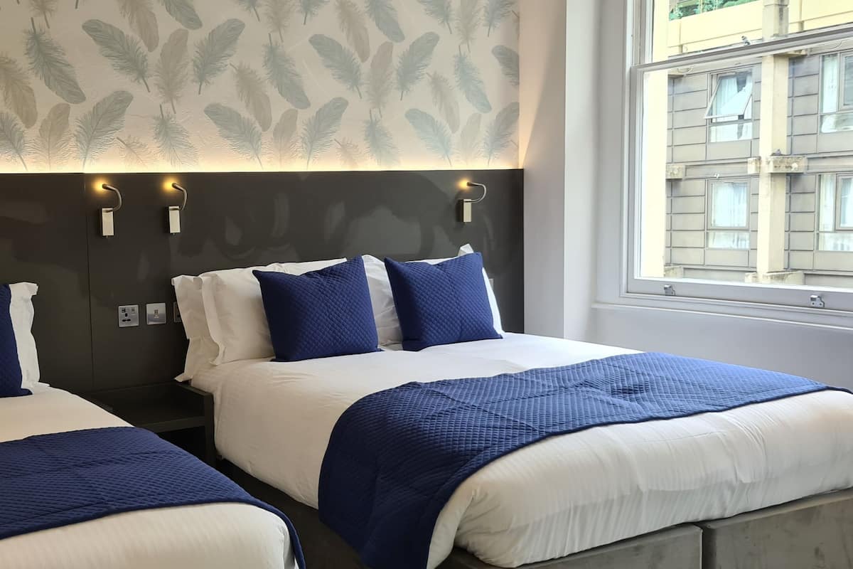 Quarto de hotel com duas camas, cada uma com roupa de cama azul e branca, e uma parede com padrão de folhas.