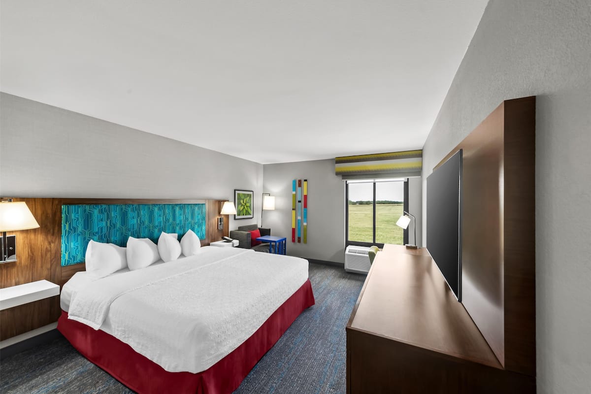 Quarto de hotel com cama grande, escrivaninha com TV de tela plana, janela com vista para o exterior e parede com obra de arte abstrata colorida.