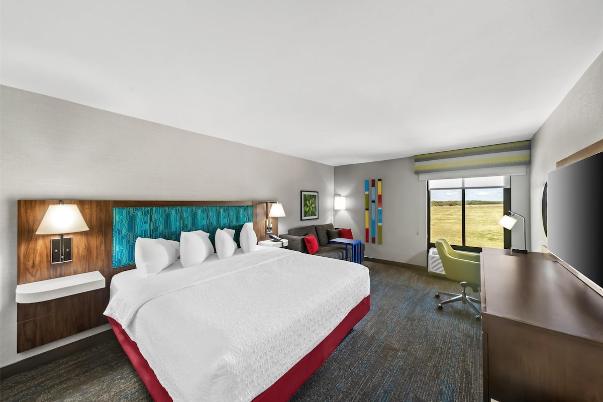 Quarto de hotel com uma cama grande, uma escrivaninha, uma cadeira, uma mesa de cabeceira, uma luminária e uma janela com vista para o exterior.