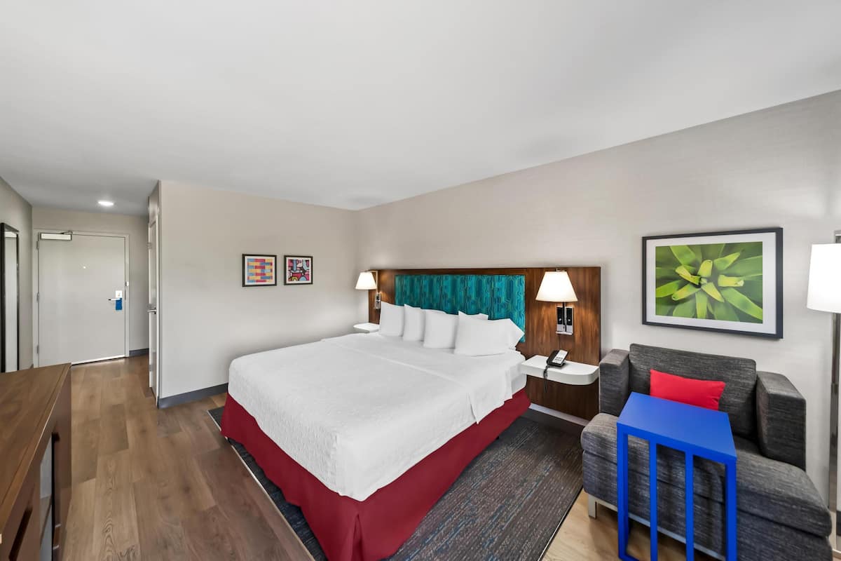Quarto de hotel com cama grande, escrivaninha, sofá e mesa de cabeceira com abajur.