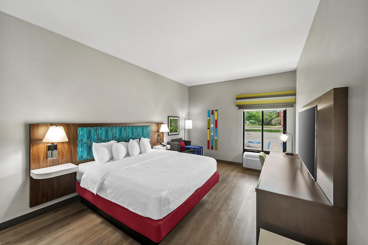 Quarto de hotel moderno com uma cama grande, uma mesa de trabalho, uma TV e uma janela com vista para o verde.