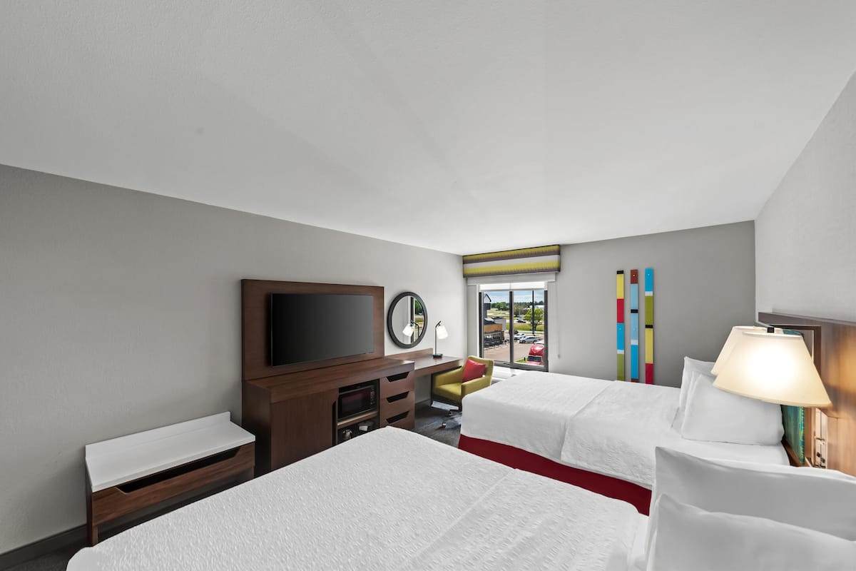 Quarto de hotel com duas camas, televisão, escrivaninha com cadeira, janela com vista e luminária.