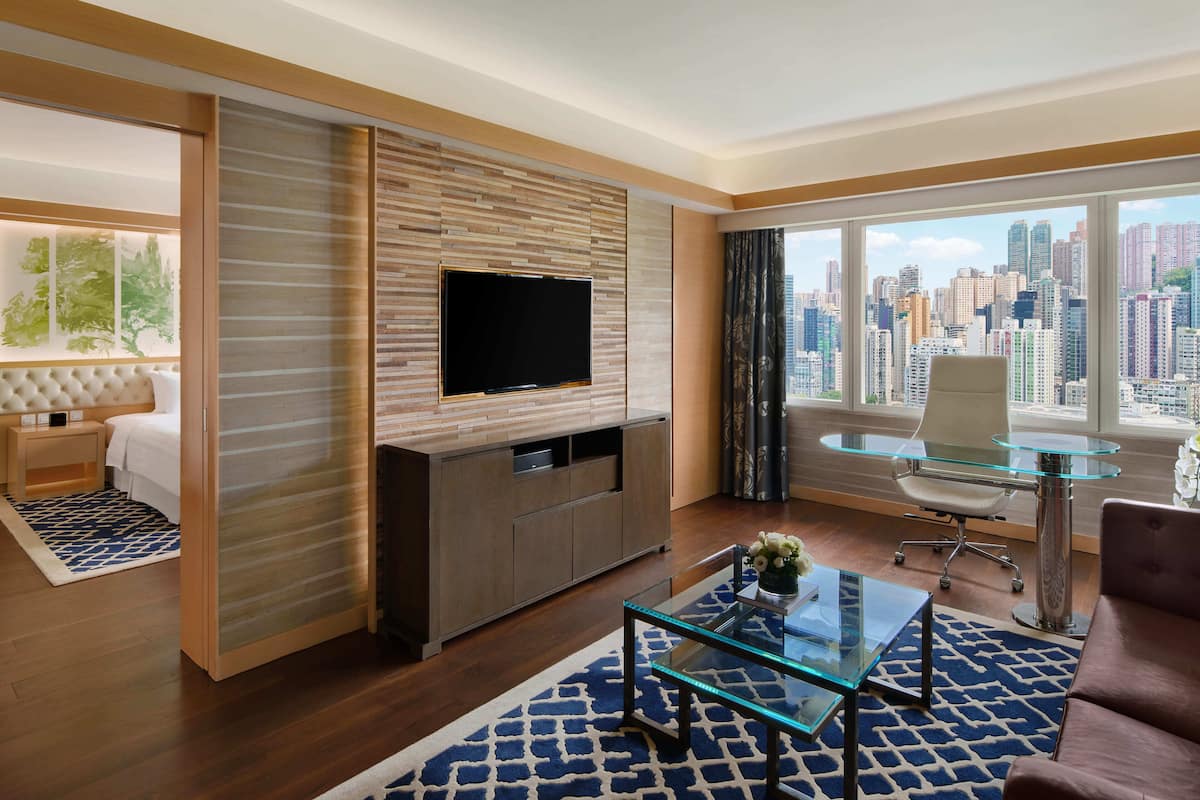 Quarto de hotel moderno com uma TV grande, uma mesa de jantar de vidro, um sofá e vista para o horizonte da cidade.