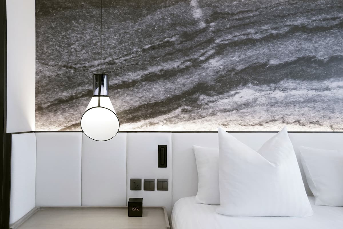 Quarto moderno com parede de mármore, luminária pendente, cama com roupa de cama branca e mesa de cabeceira com relógio e tomadas.