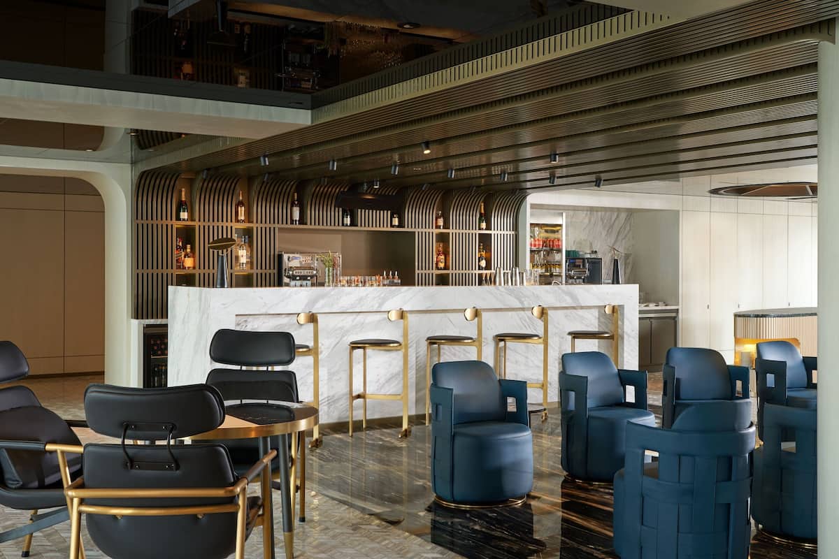 Uma área de bar moderna com bancada de mármore, banquetas pretas e azuis e teto alto com ripas de madeira.