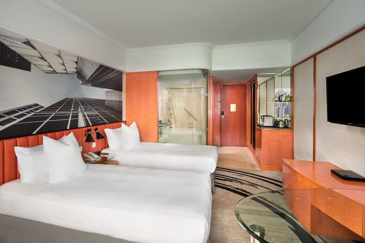 Quarto de hotel com duas camas, televisão, frigobar e banheiro com chuveiro de vidro.