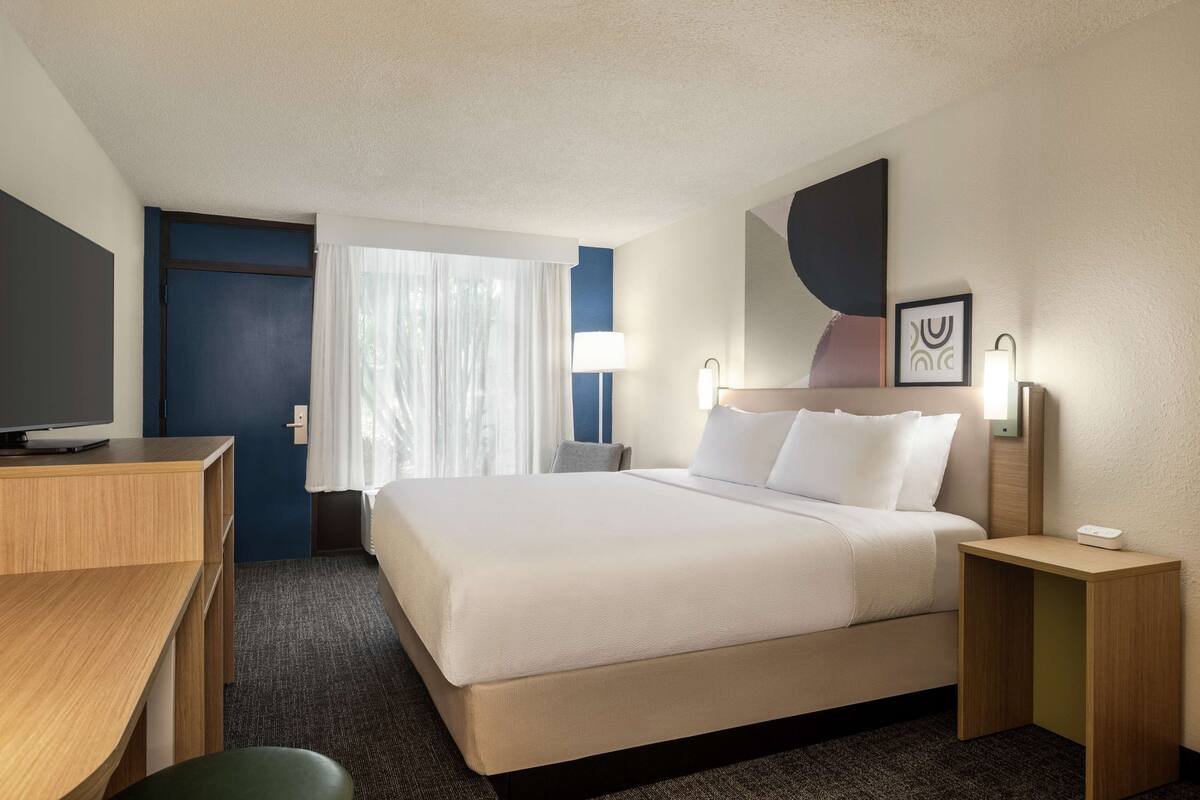 Quarto de hotel com cama, escrivaninha, cadeira, mesa de cabeceira e luminária.