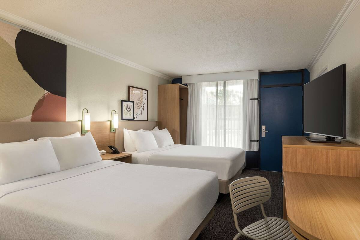 Quarto de hotel com duas camas, uma escrivaninha, uma cadeira, uma televisão, mesa de cabeceira com telefone e uma janela com cortinas.