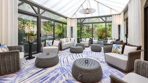 Uma sala de sol com móveis de vime, um tapete estampado e amplas janelas com vista para um jardim.