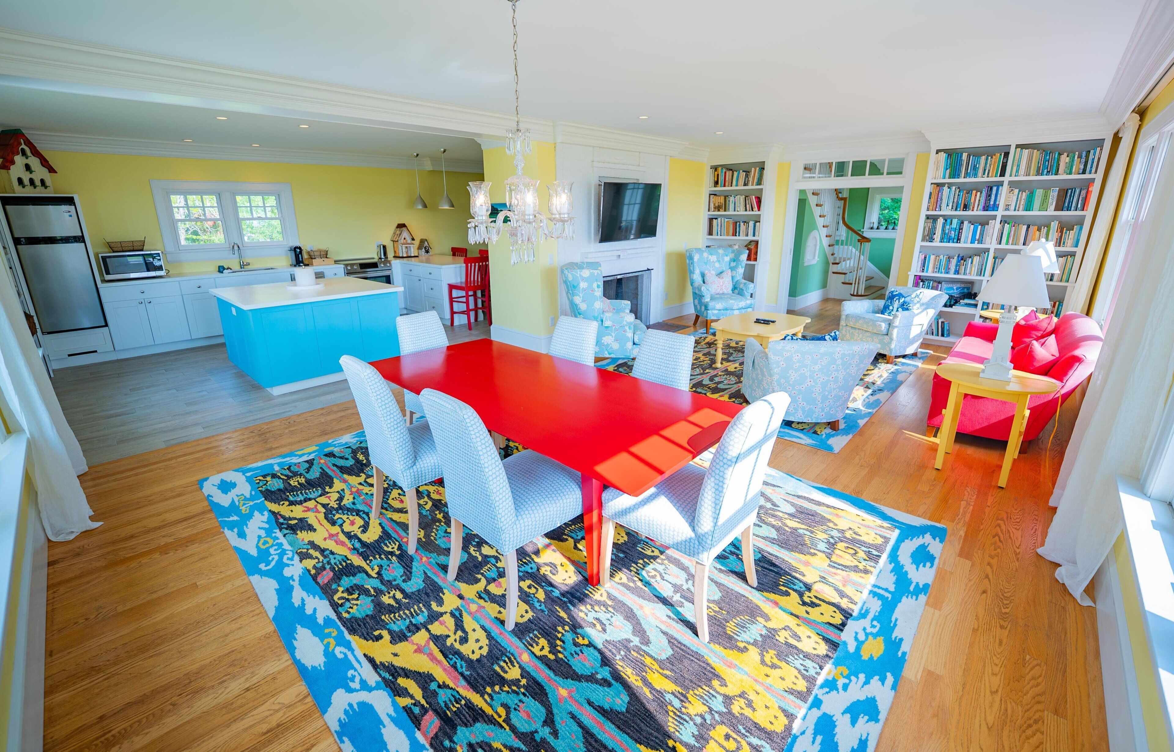 Ampla sala de estar com uma mesa de jantar vermelha, tapete estampado em azul e uma cozinha ao fundo.