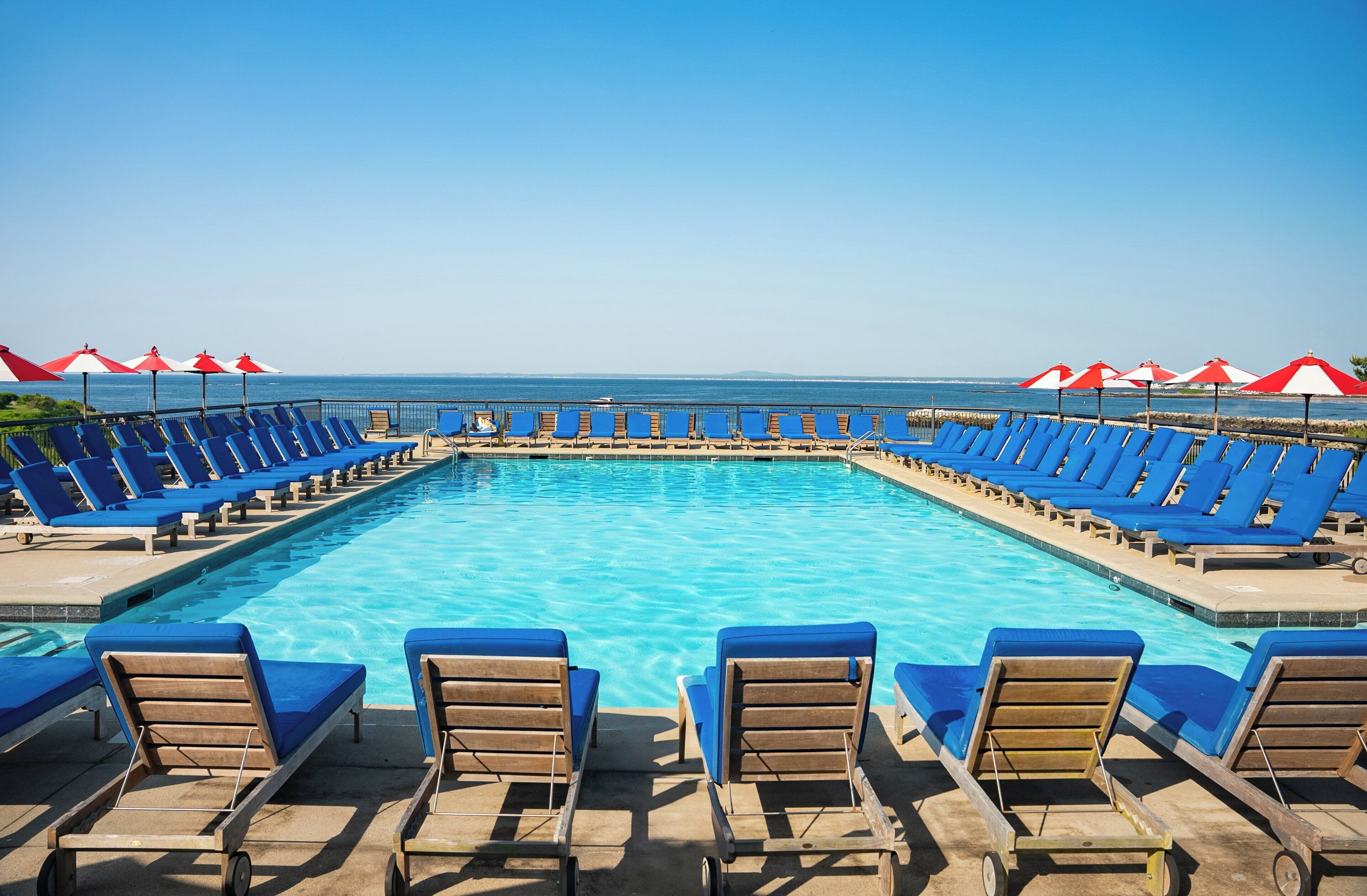 Área de piscina com espreguiçadeiras azuis e guarda-sóis vermelhos, com vista para o mar azul e límpido.