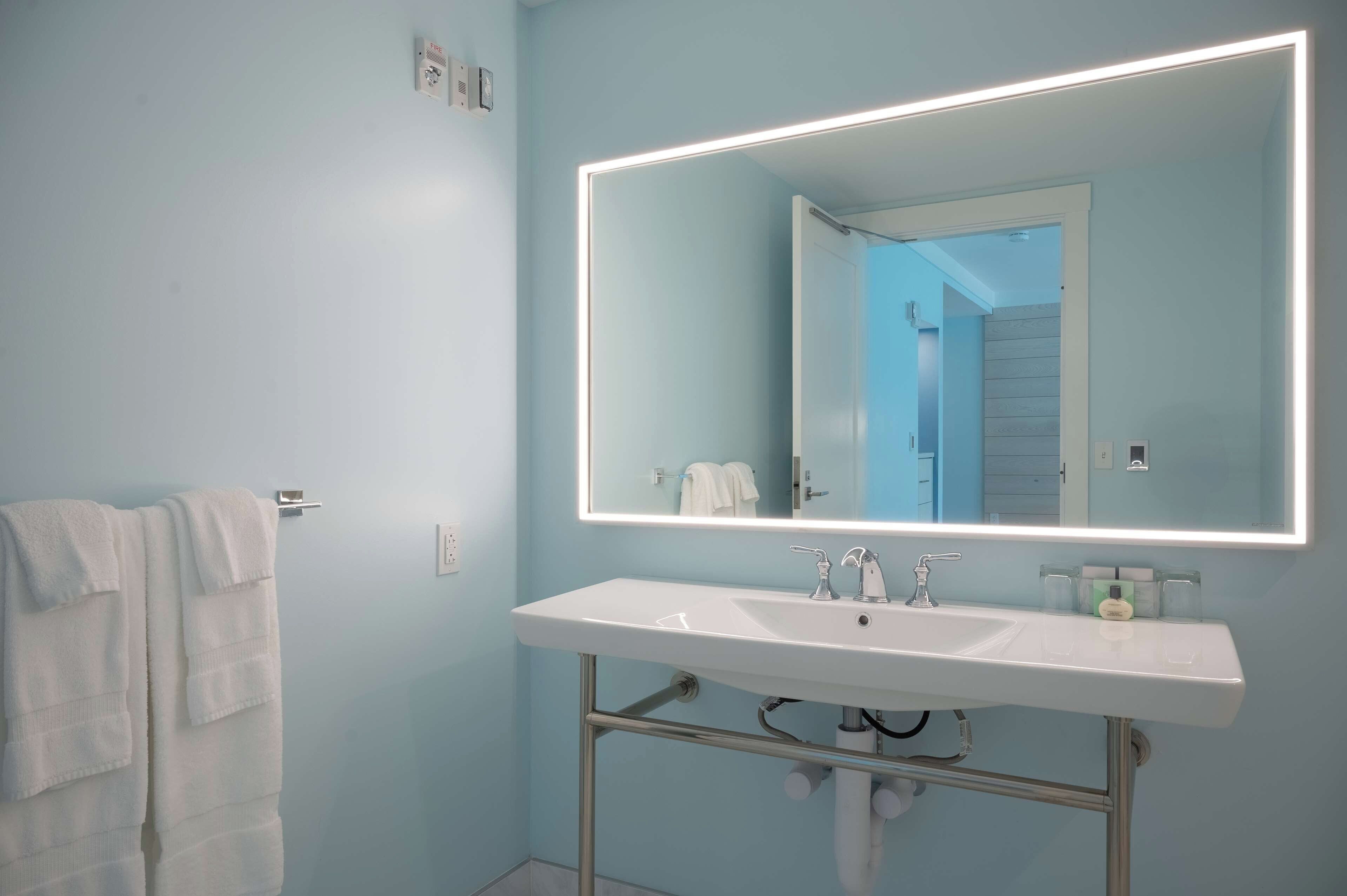 Uma casa de banho com uma pia branca, um espelho grande com iluminação LED e um porta-toalhas com toalhas dobradas.