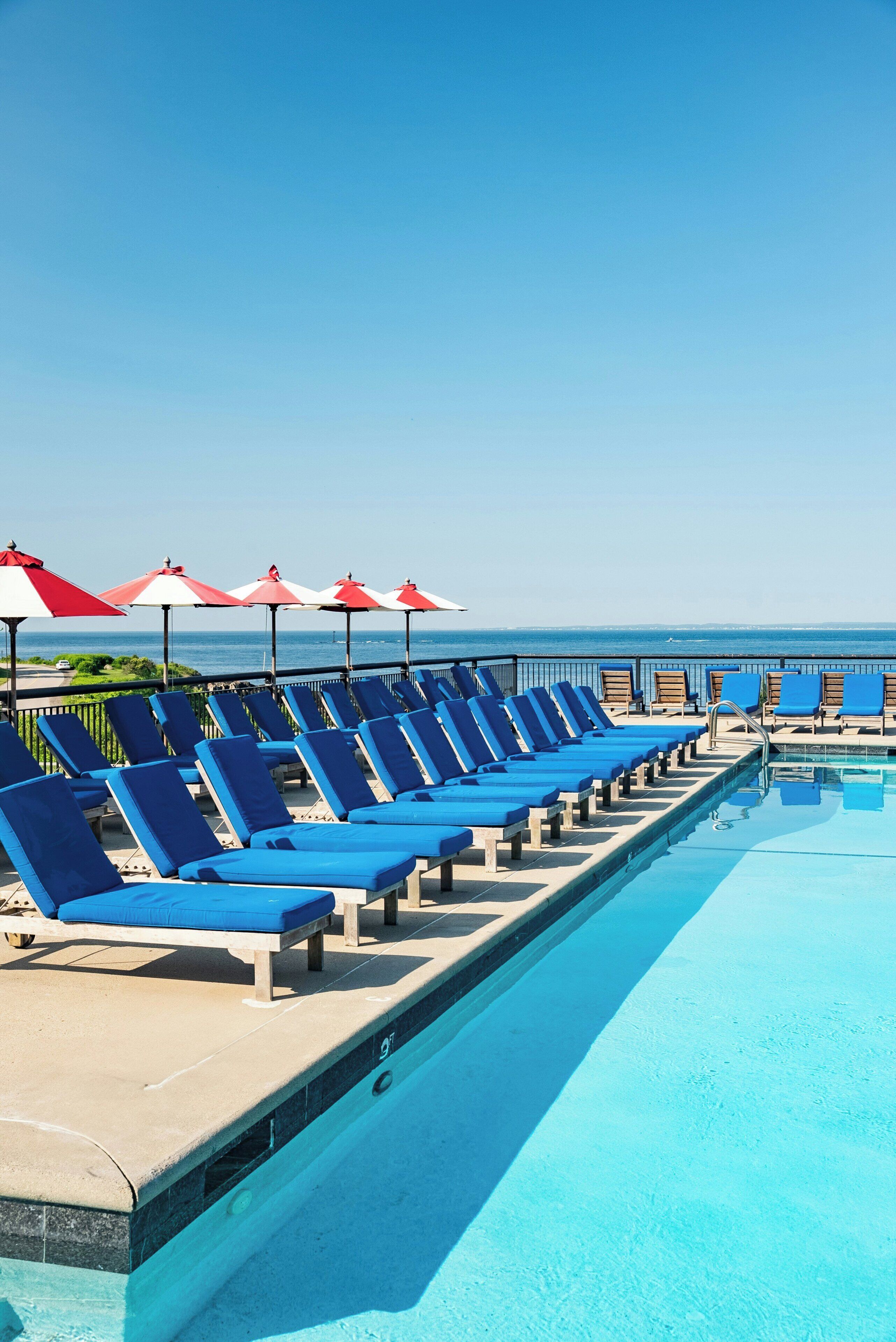 Área à beira da piscina com cadeiras de descanso azuis e guarda-sóis vermelhos, céu claro e vista para o oceano.