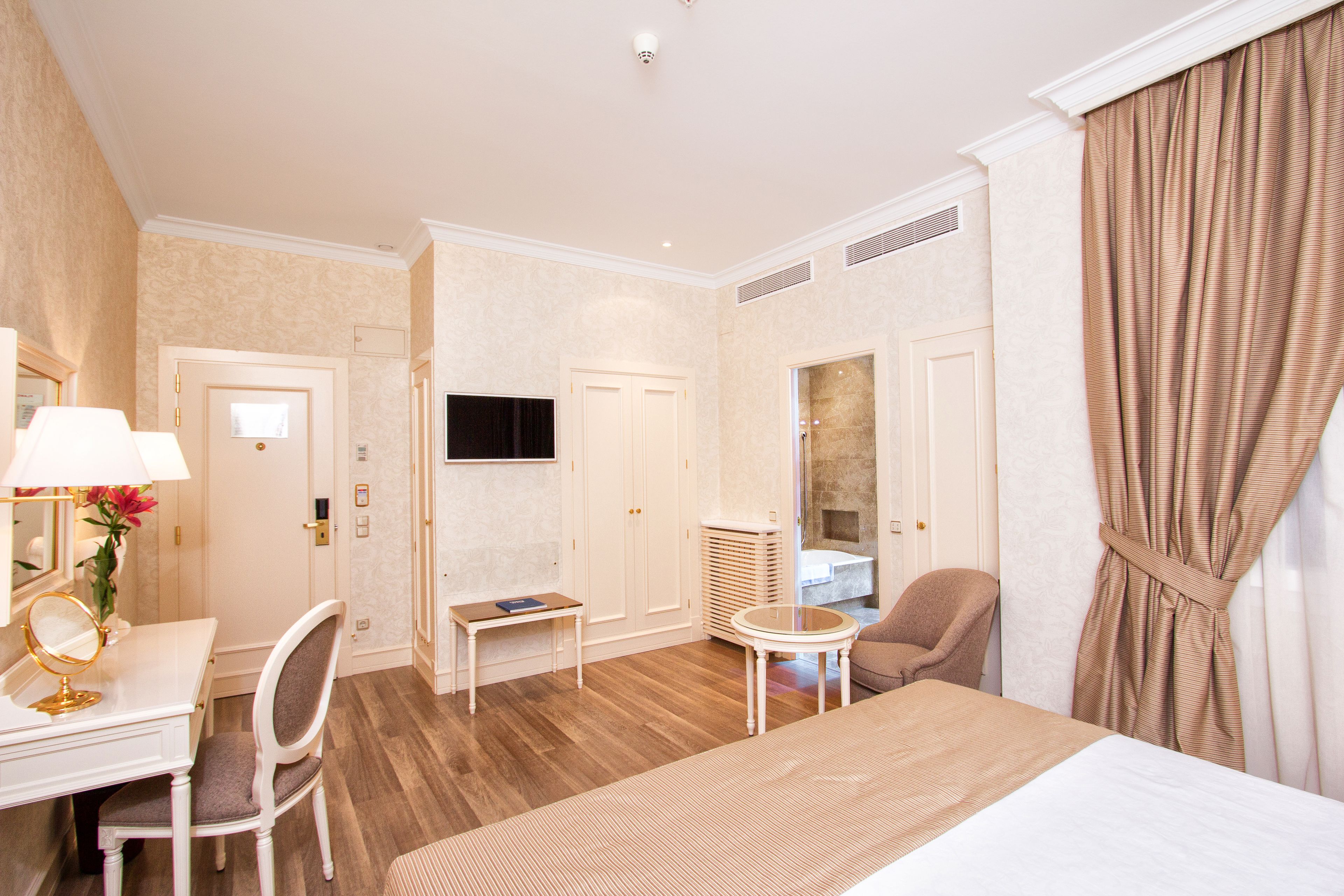 Quarto de hotel com cama, escrivaninha com cadeira, TV na parede e banheiro com chuveiro e vaso sanitário.