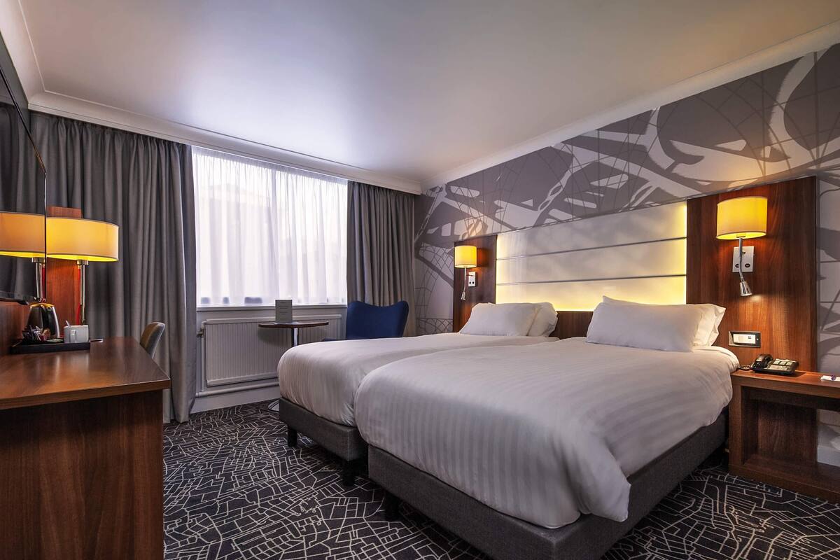 Quarto de hotel com uma cama grande, duas mesas de cabeceira com luminárias, uma escrivaninha com cadeira, uma mesinha e uma janela com cortinas.