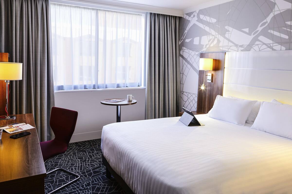 Quarto de hotel com uma cama grande, uma mesa com luminária, uma cadeira, uma mesinha com uma revista e uma janela com cortinas.