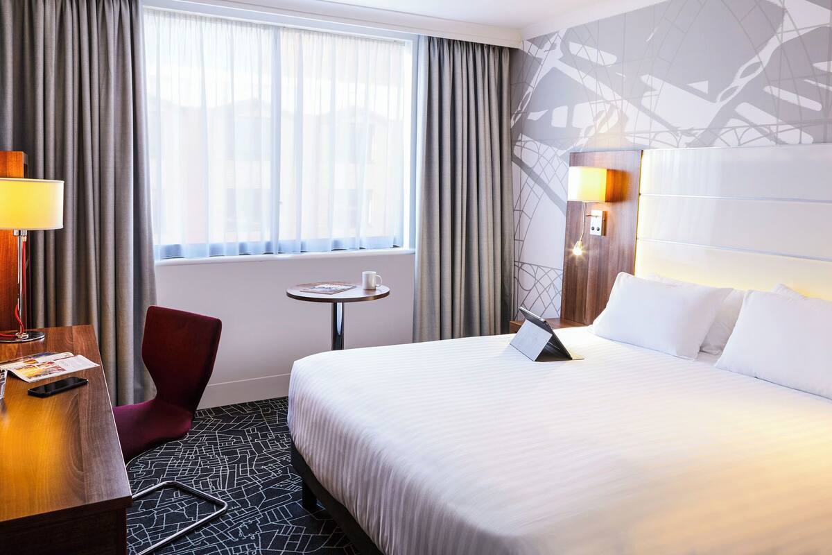 Quarto de hotel com uma cama grande, uma escrivaninha com luminária, uma cadeira, uma mesinha e uma janela com cortinas.