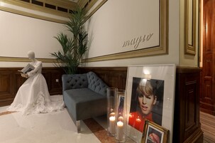 Uma área de recepção com um manequim vestindo um vestido branco, um sofá cinzento, vasos altos com velas e fotografias emolduradas na parede.