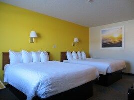 Quarto de hotel com duas camas, cada uma com roupa de cama e almofadas brancas, em frente a uma parede amarela. Há duas luminárias fixadas na parede e um quadro com uma imagem de um pôr do sol.
