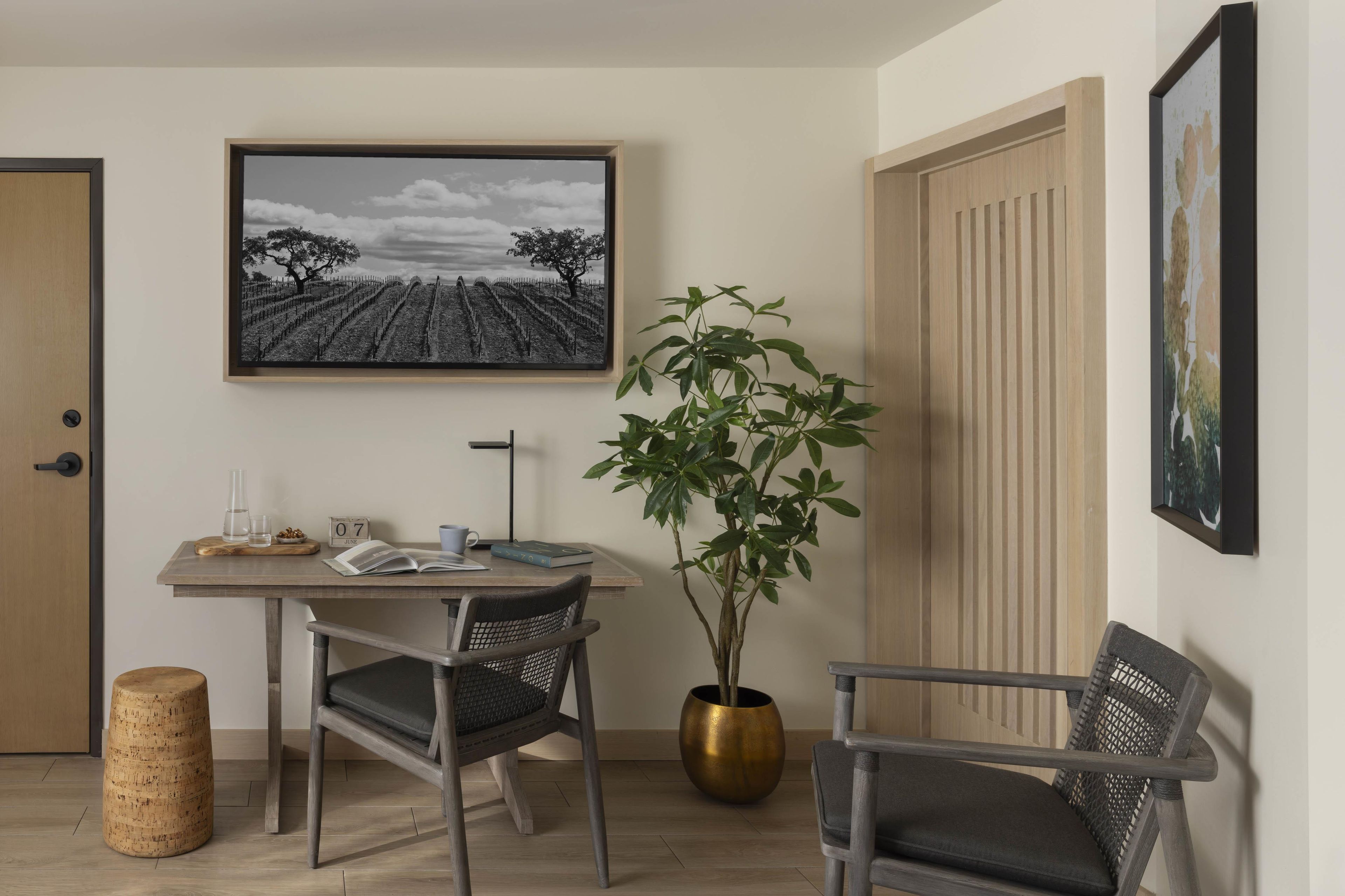 Quarto com escrivaninha de madeira, duas cadeiras, um vaso de planta, um quadro na parede e uma porta.
