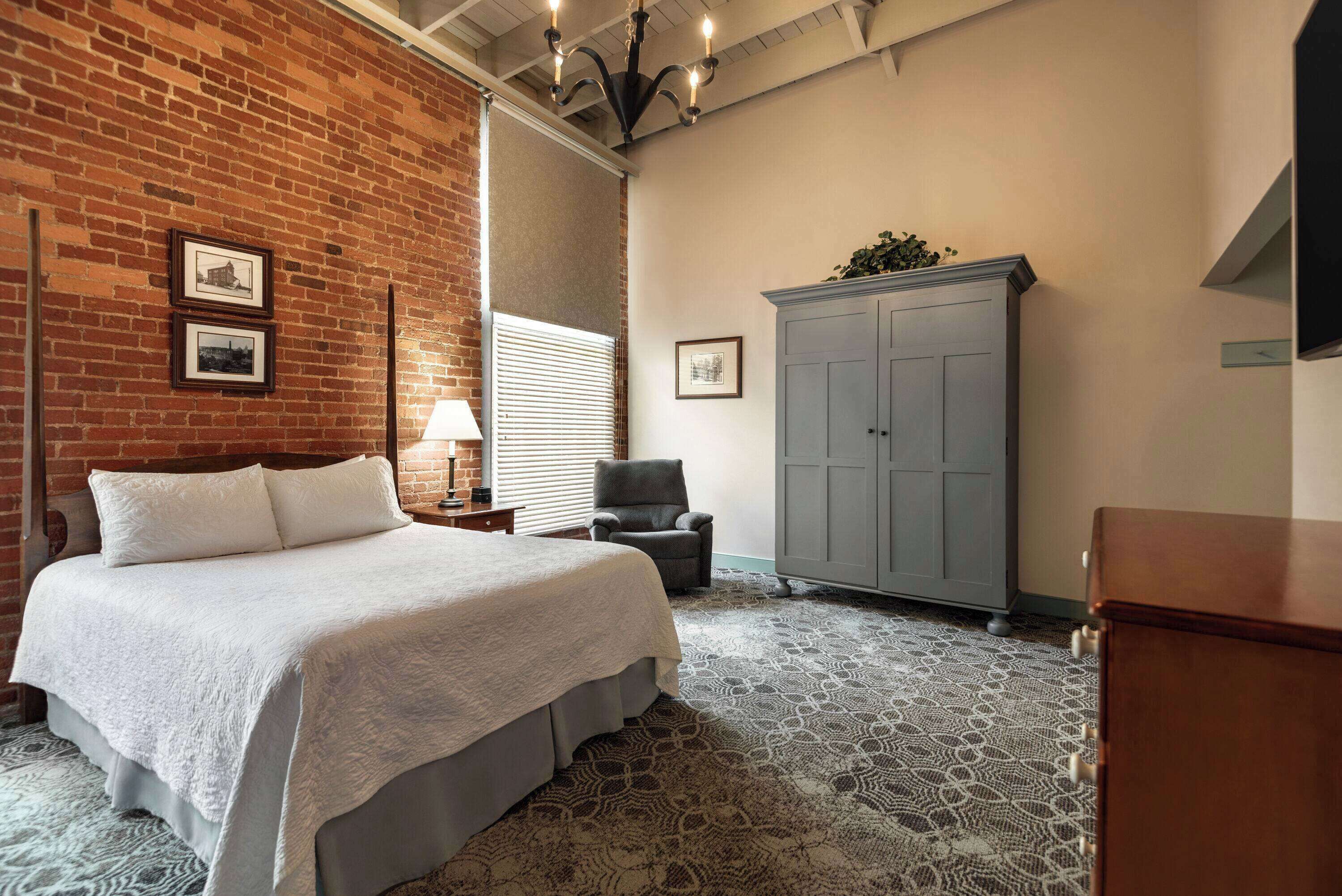 Quarto com parede de tijolos, uma cama grande, um guarda-roupa cinza e um candelabro.
