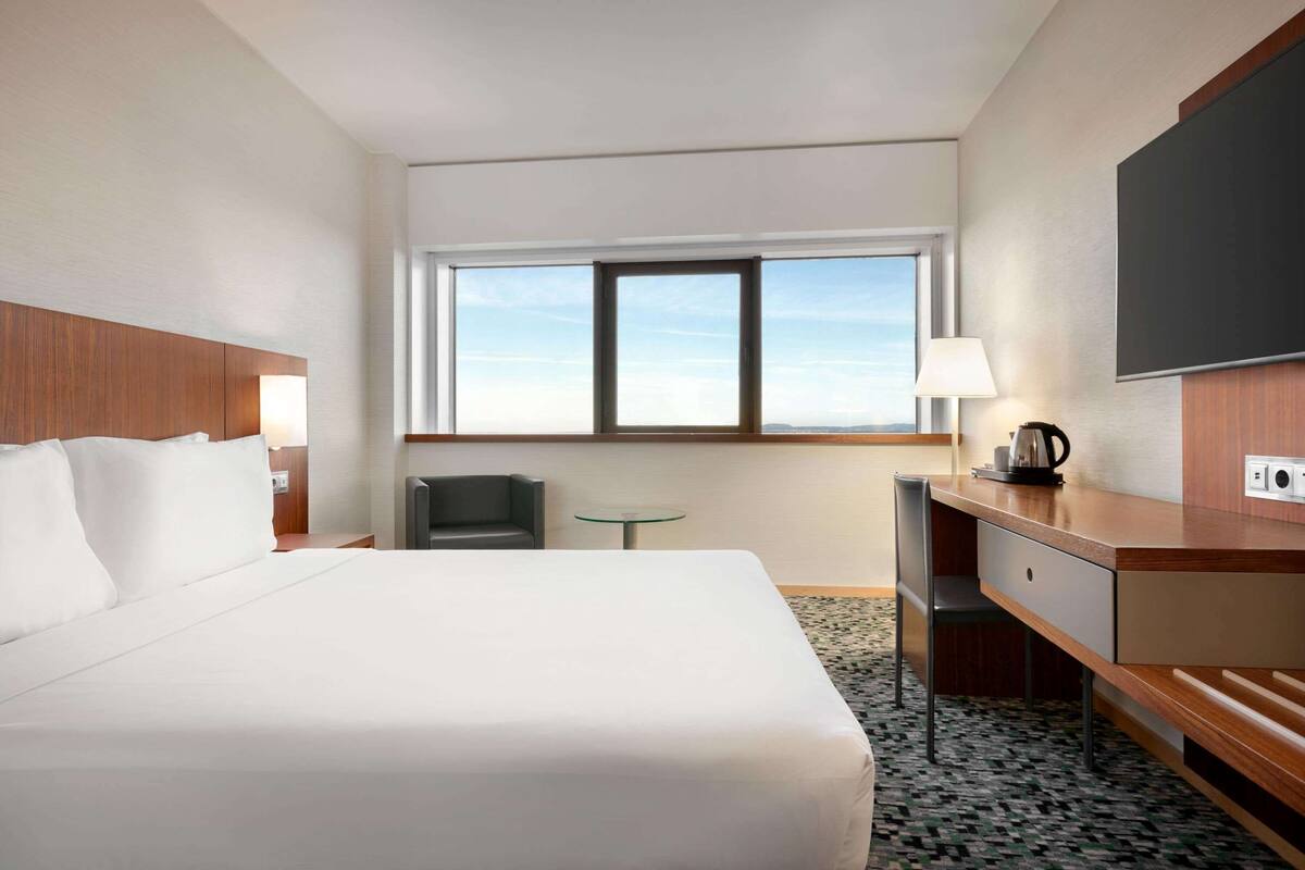 Quarto de hotel com uma cama grande, uma escrivaninha com abajur, televisão e janela com vista para o céu.