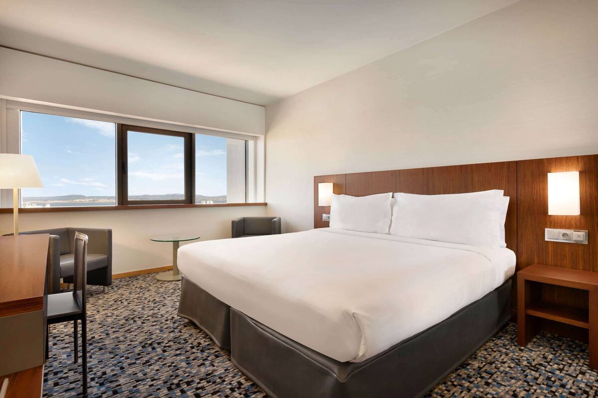 Quarto de hotel com uma cama grande, uma escrivaninha com cadeira, uma pequena mesa e uma mesinha de cabeceira. Há uma janela com vista para o céu e a paisagem distante.