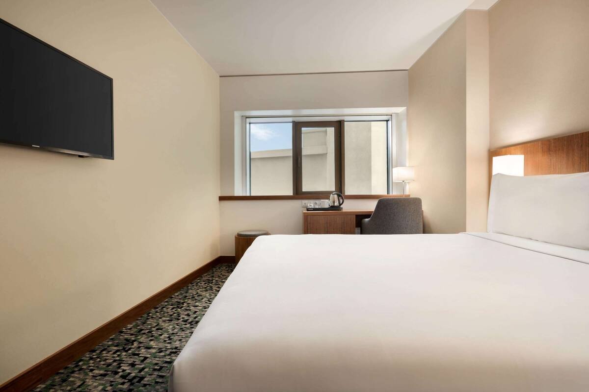 Quarto de hotel com uma cama grande, uma escrivaninha com cadeira, televisão de tela plana na parede e uma janela com vista para o exterior.