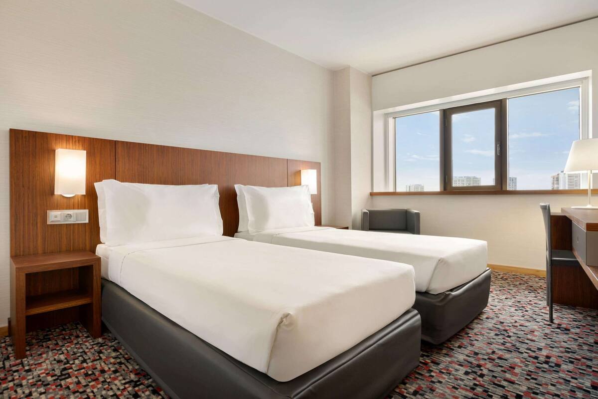 Quarto de hotel com duas camas, uma escrivaninha e janela com vista para a cidade.