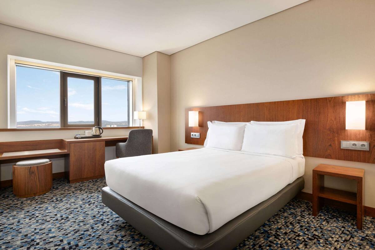 Quarto de hotel com uma cama grande, uma escrivaninha, uma cadeira, um banquinho pequeno, uma mesa de cabeceira e uma janela com vista para a paisagem.