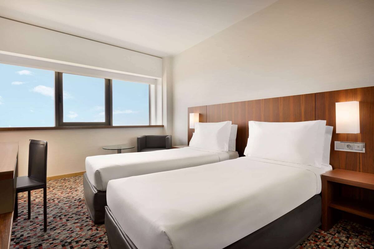 Quarto de hotel com duas camas, uma escrivaninha, uma cadeira e uma janela com vista.