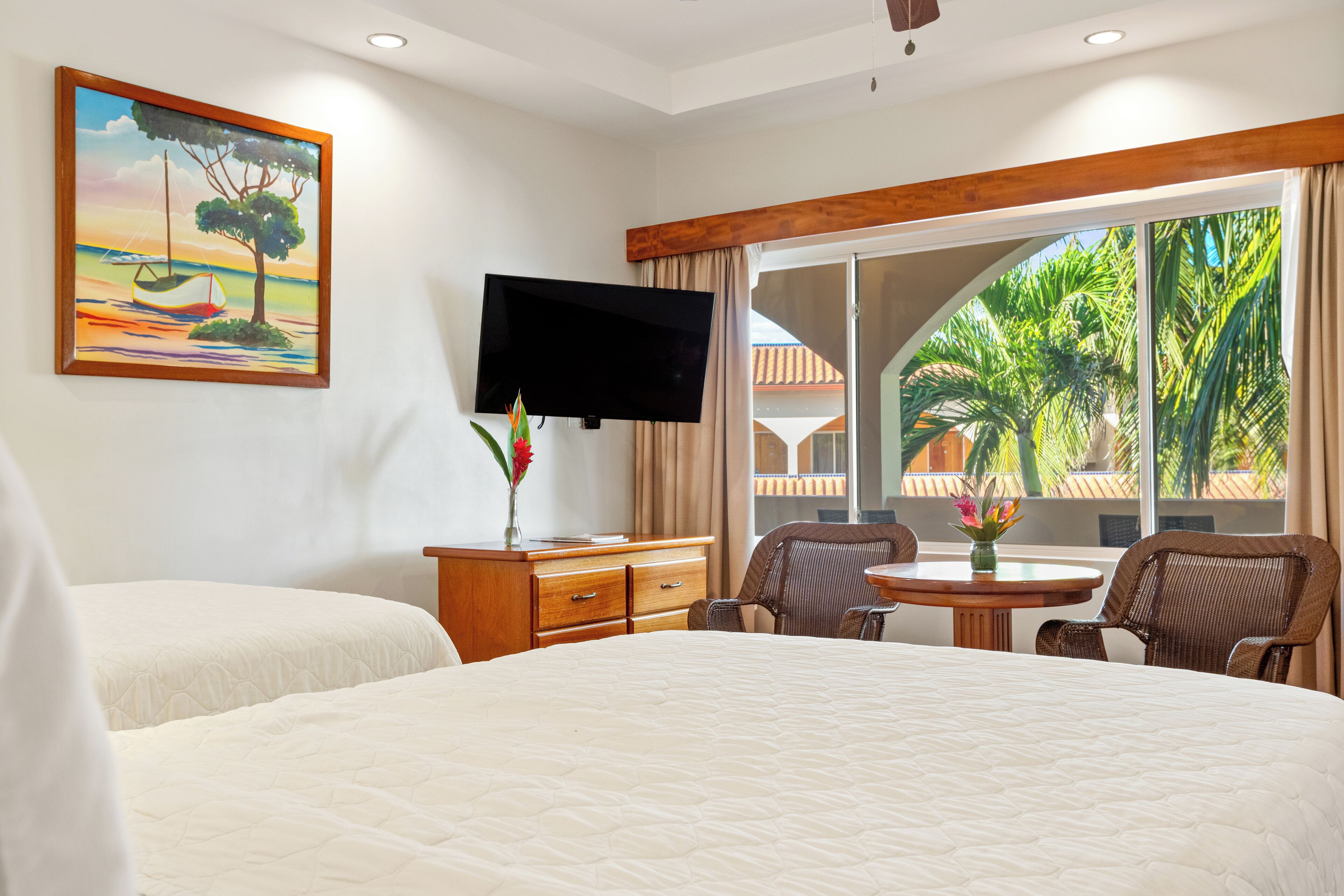 Quarto de hotel com duas camas, televisão, mesa de jantar e cadeiras, e uma grande janela com vista para palmeiras.