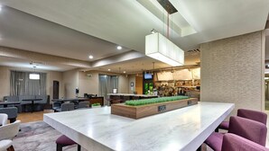 Um hall de hotel moderno com bar central, áreas de estar e um amplo balcão de recepção em mármore.