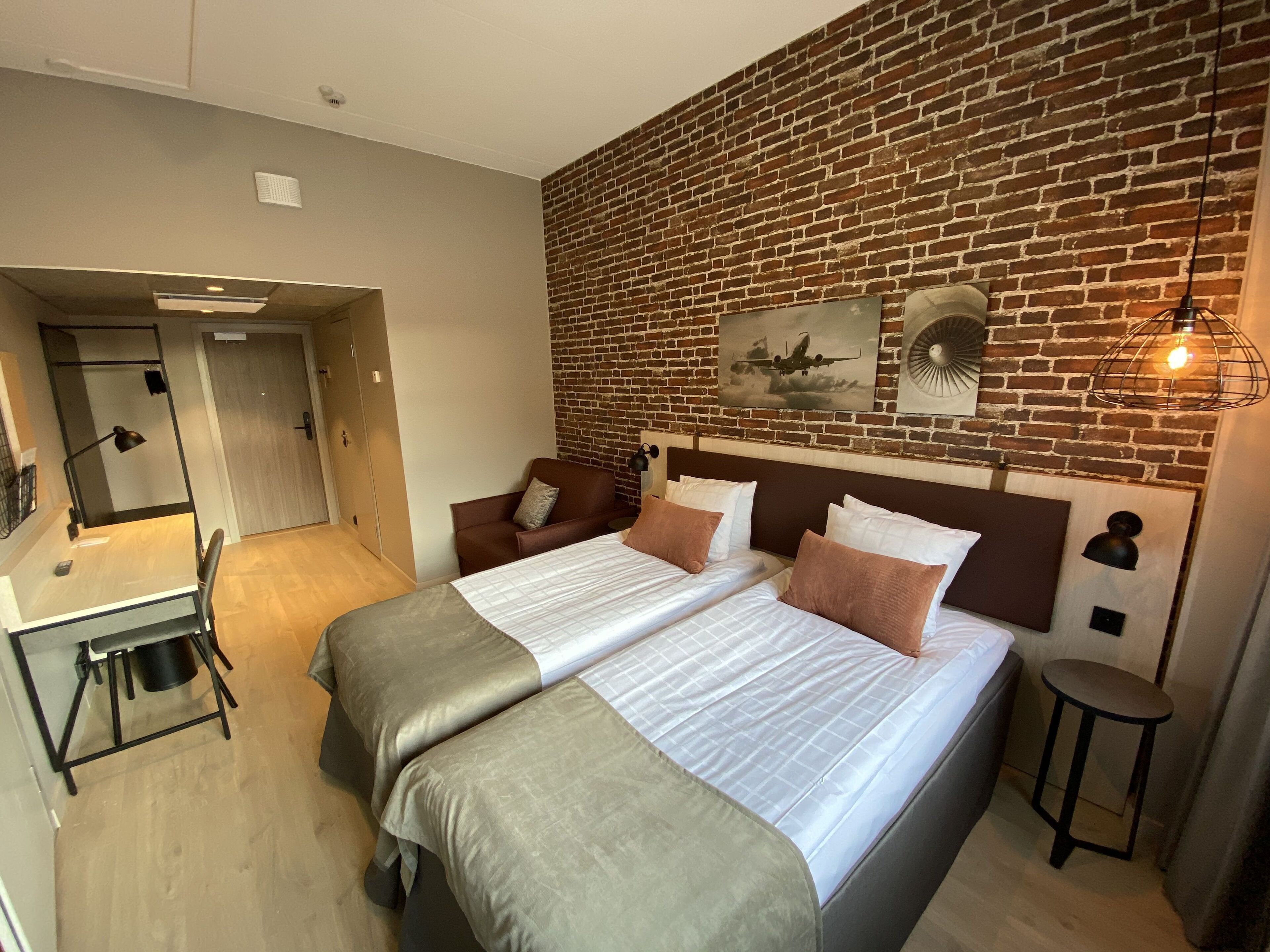 Quarto de hotel com parede de tijolos, cama com roupa de cama branca e castanha, escrivaninha com cadeira e um banquinho pequeno.
