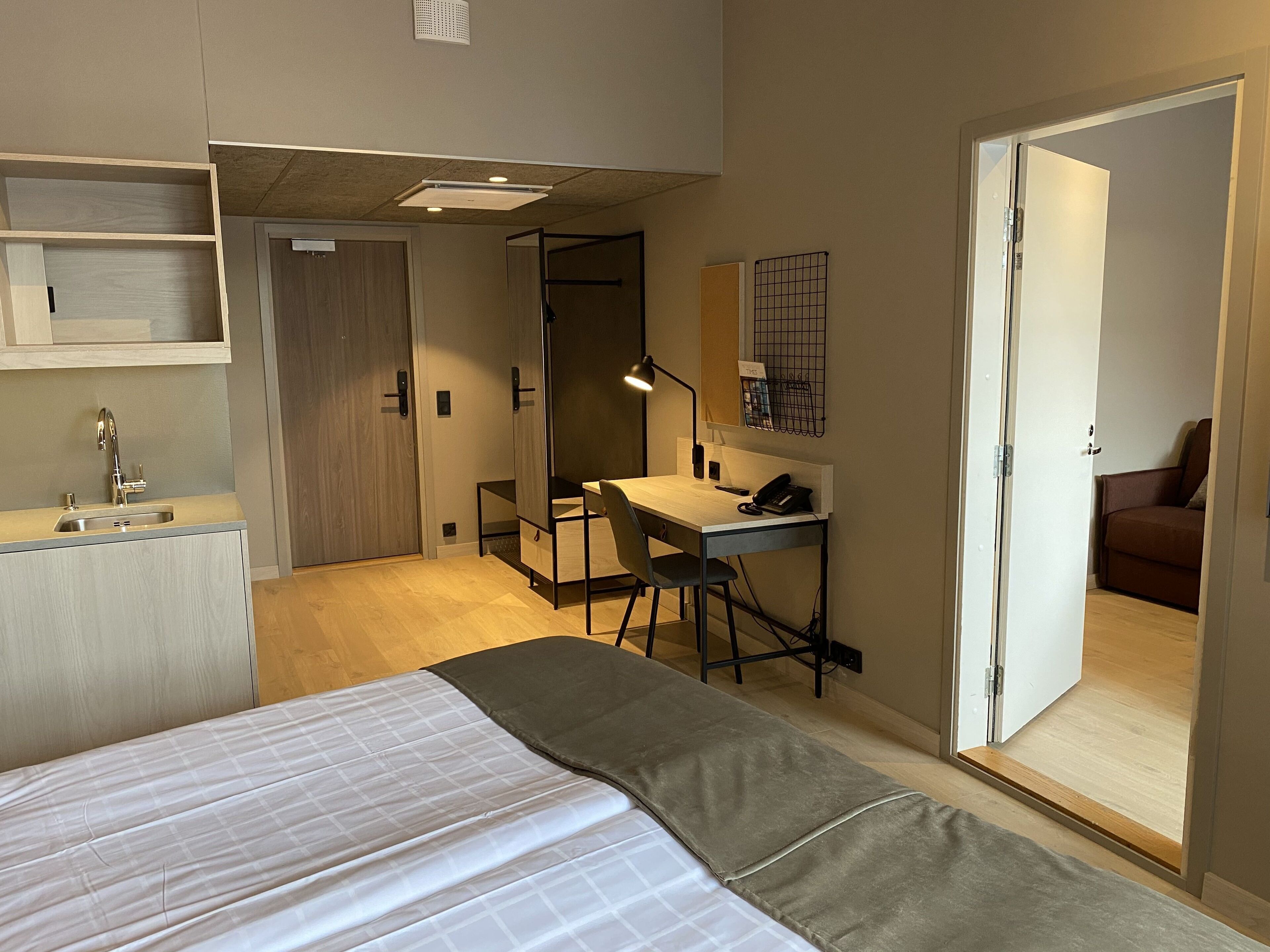 Quarto de hotel compacto com cama, escrivaninha e cadeira, pequeno espaço de cozinha e porta que dá para outro quarto.