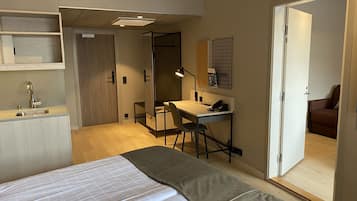 Quarto de hotel compacto com cama, escrivaninha e cadeira, pequeno espaço de cozinha e porta que dá para outro quarto.