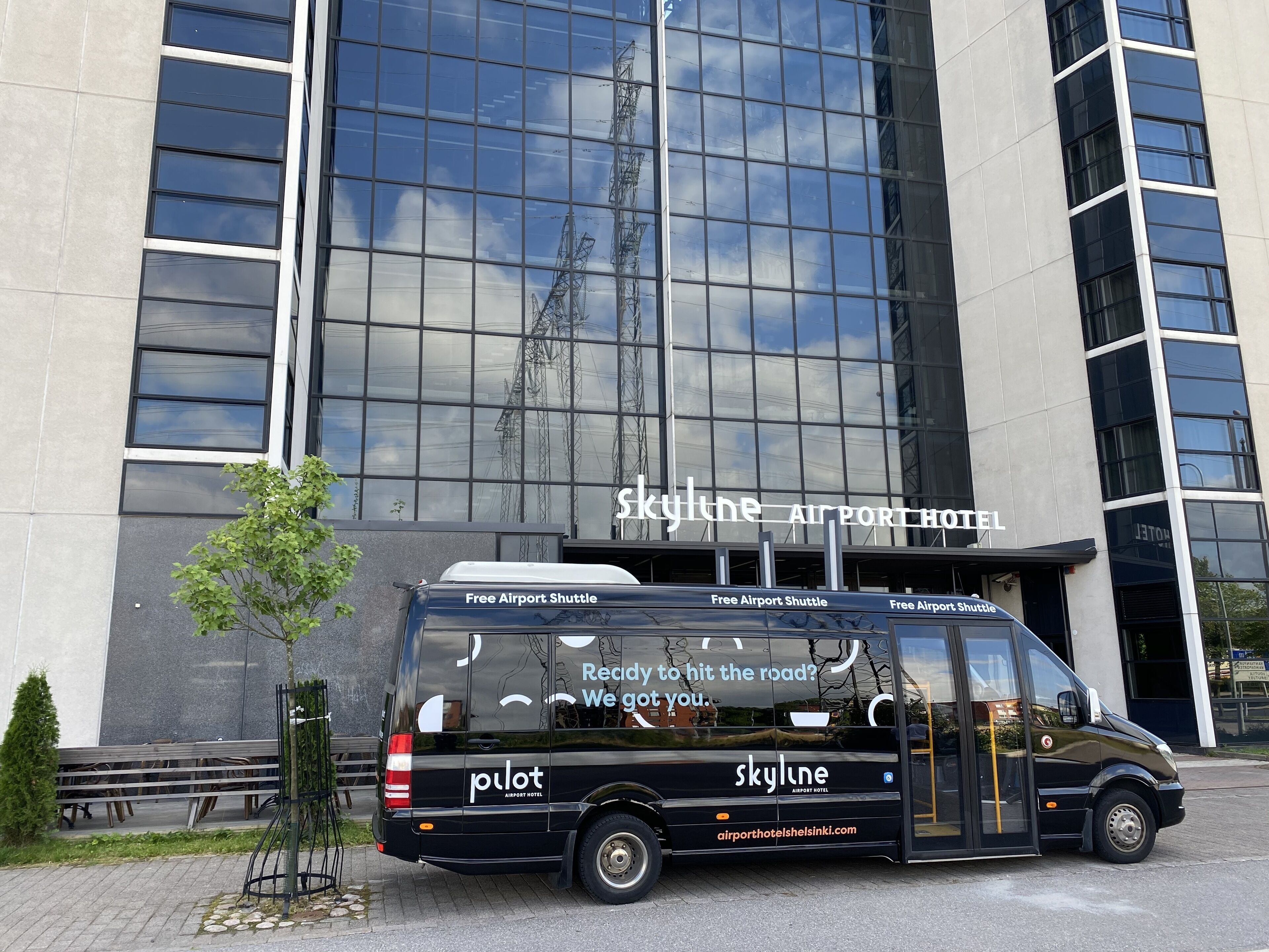Um ônibus executivo preto estacionado em frente ao Skyline Airport Hotel, com uma placa indicando que oferece serviço de transporte gratuito para o aeroporto.