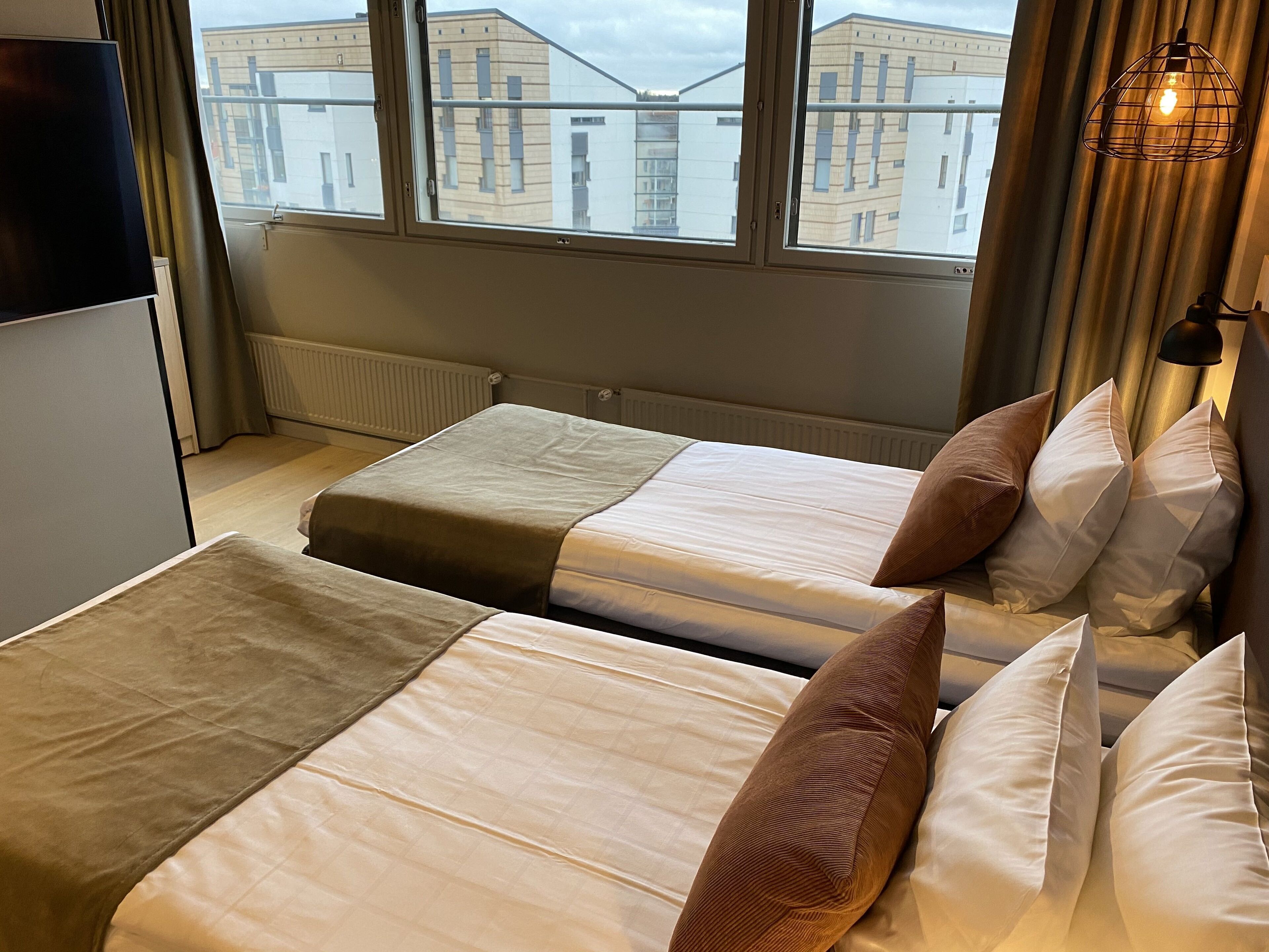 Quarto de hotel com duas camas, uma televisão, um radiador e vista para edifícios pela janela.