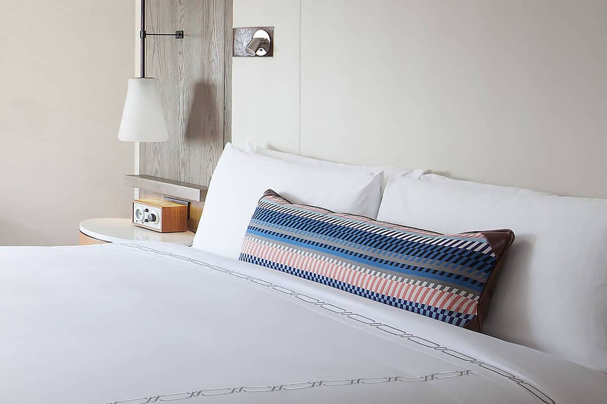 Uma cama bem arrumada com lençóis brancos e almofada estampada, uma mesa de cabeceira com um rádio e um abajur fixado na parede.