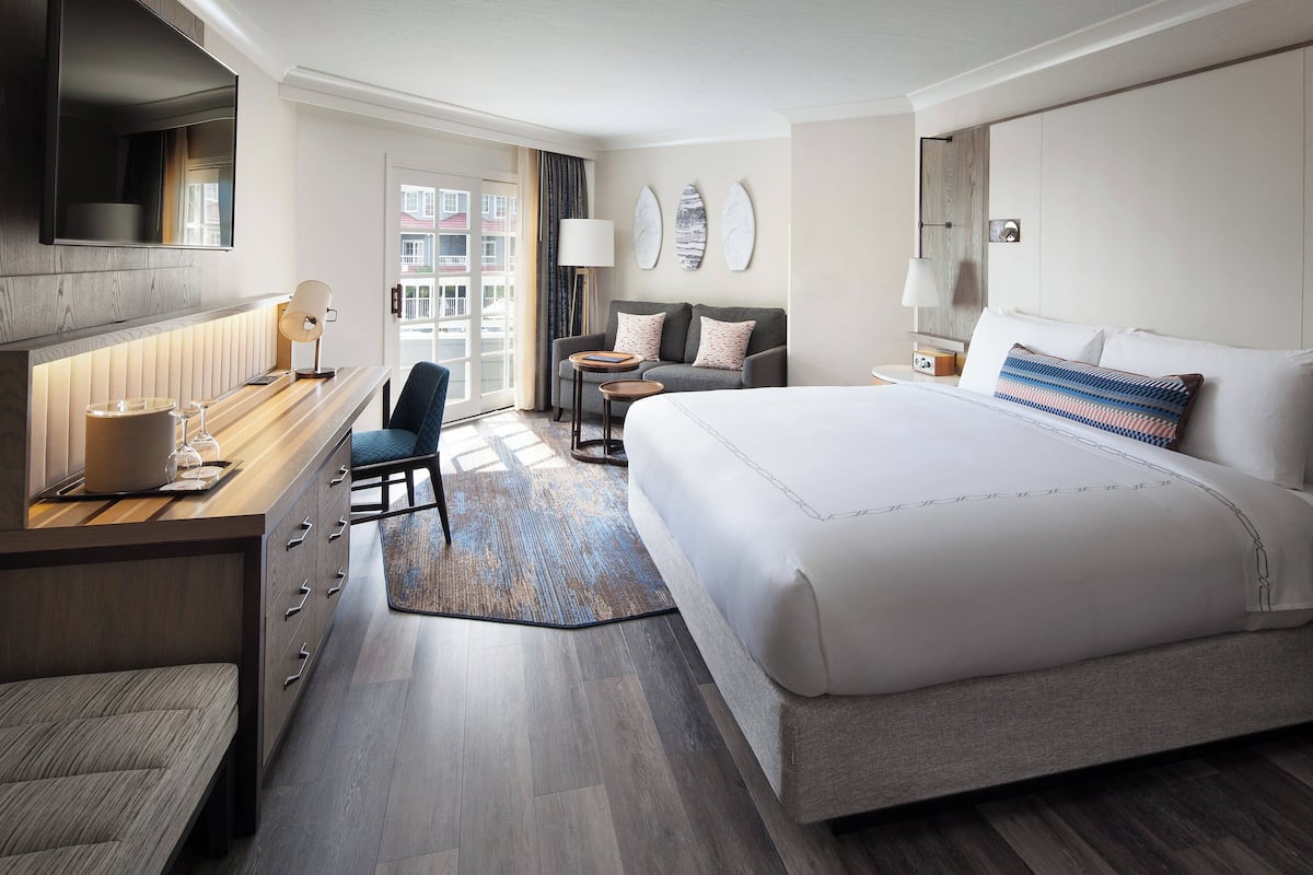 Quarto de hotel com uma cama grande, uma escrivaninha com luminária, uma cadeira e uma mesinha.